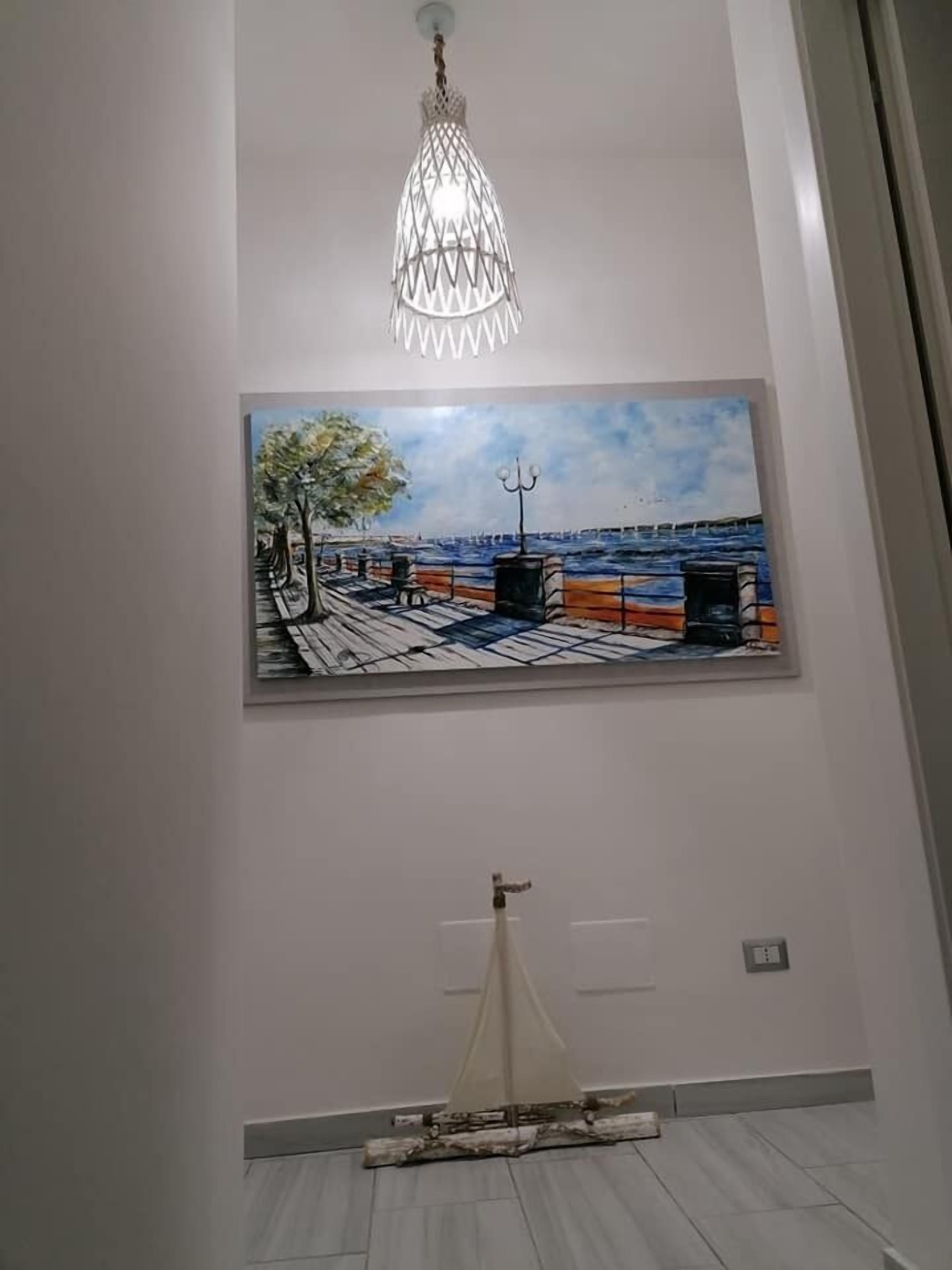 Pintura de um cais com uma árvore e um candelabro, uma luminária geométrica branca e um modelo de veleiro no chão.