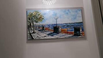 Pintura de um cais com uma árvore e um candelabro, uma luminária geométrica branca e um modelo de veleiro no chão.