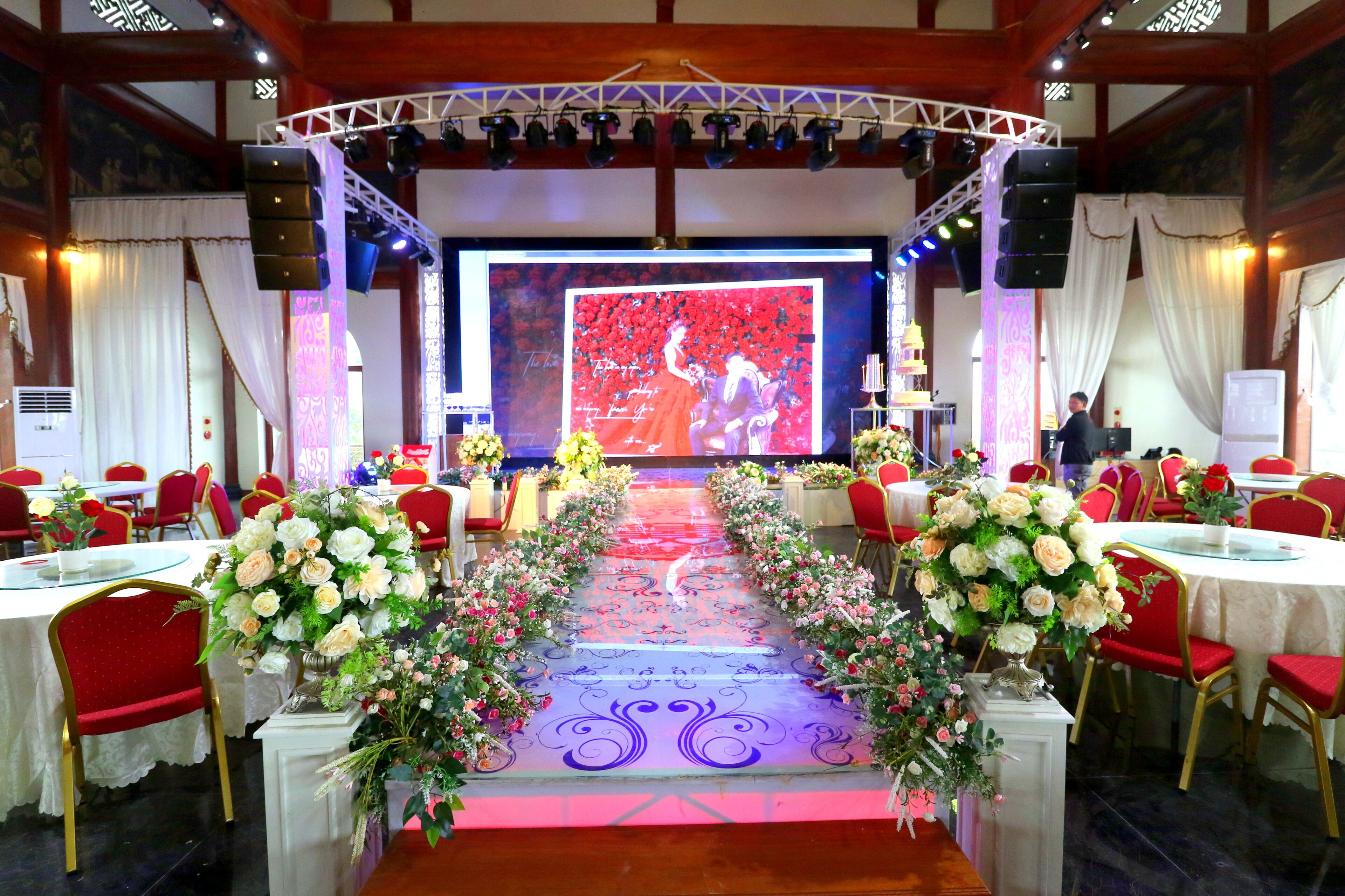 Uma sala de banquetes com palco, cadeiras vermelhas e arranjos florais.