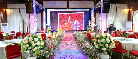 Uma sala de banquetes com palco, cadeiras vermelhas e arranjos florais.