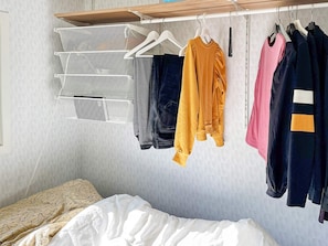 Quarto com cama, cabideiro com roupas e prateleira na parede.