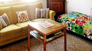 Uma sala de estar com sofá, mesa de centro, estante de livros e uma cama com colcha florida.