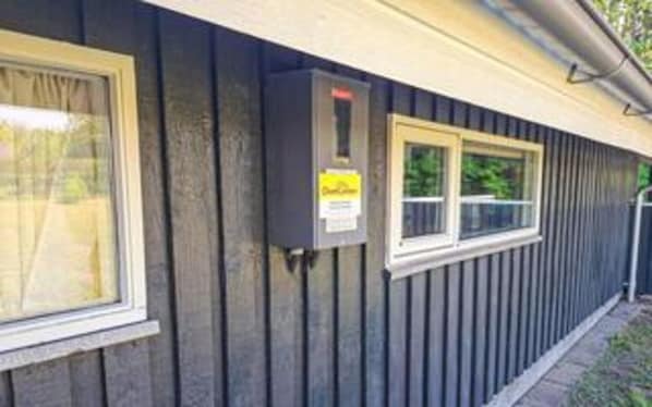 Um edifício com fachada escura, uma janela com moldura branca e um pequeno dispositivo eletrônico fixado na parede.
