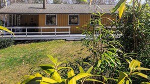 Uma casa de madeira com varanda coberta, chaminé e um gramado com plantas de bambu.