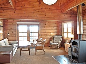 Interior de uma cabana de toras com lareira, móveis de vime e uma janela grande.