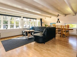 Uma sala de estar ampla com sofá de couro preto, mesa de jantar de madeira e grandes janelas.