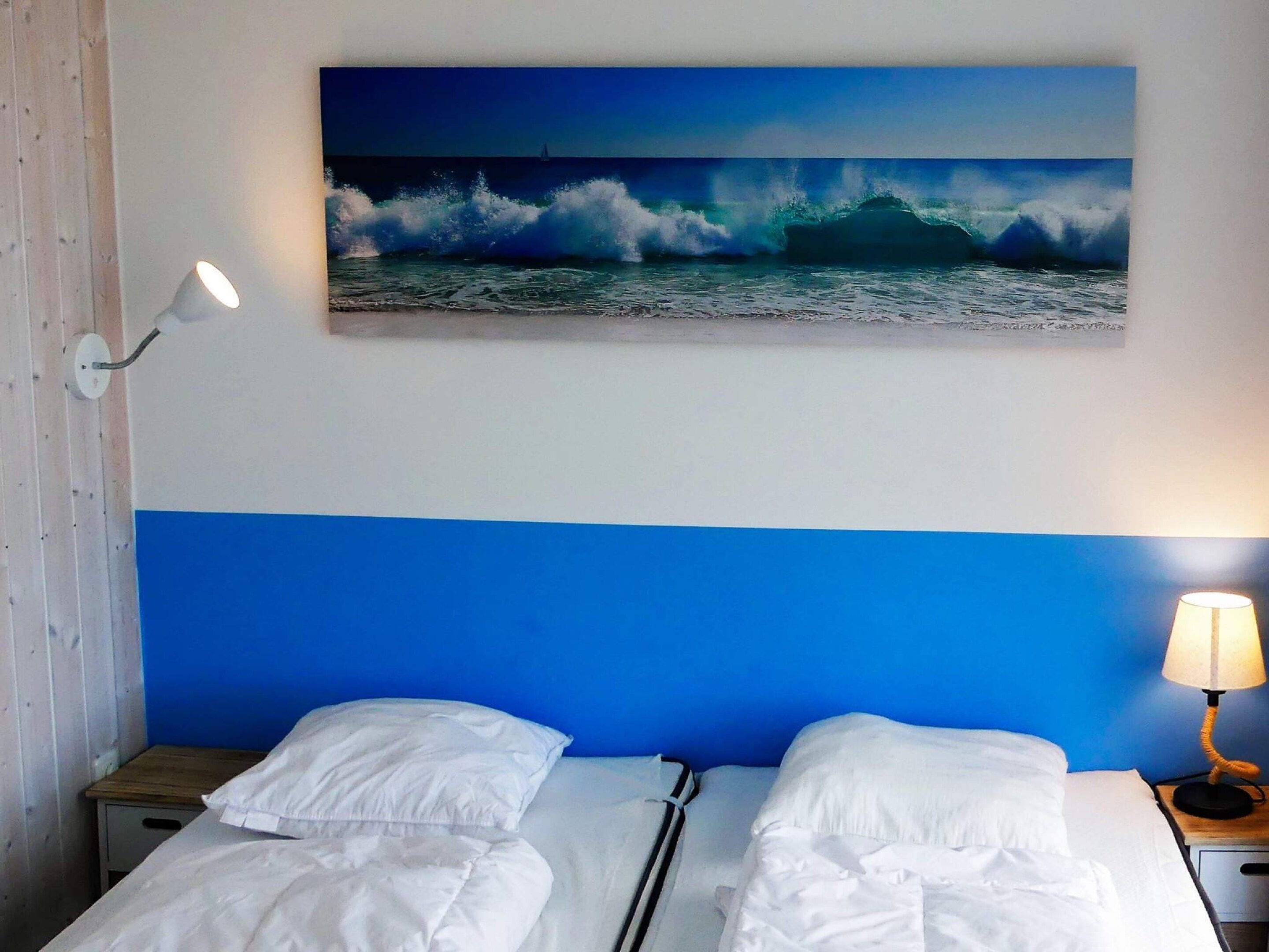 Quarto de hotel com cama, duas luminárias de cabeceira, pintura pendurada na parede retratando uma onda e mesa de cabeceira de madeira.
