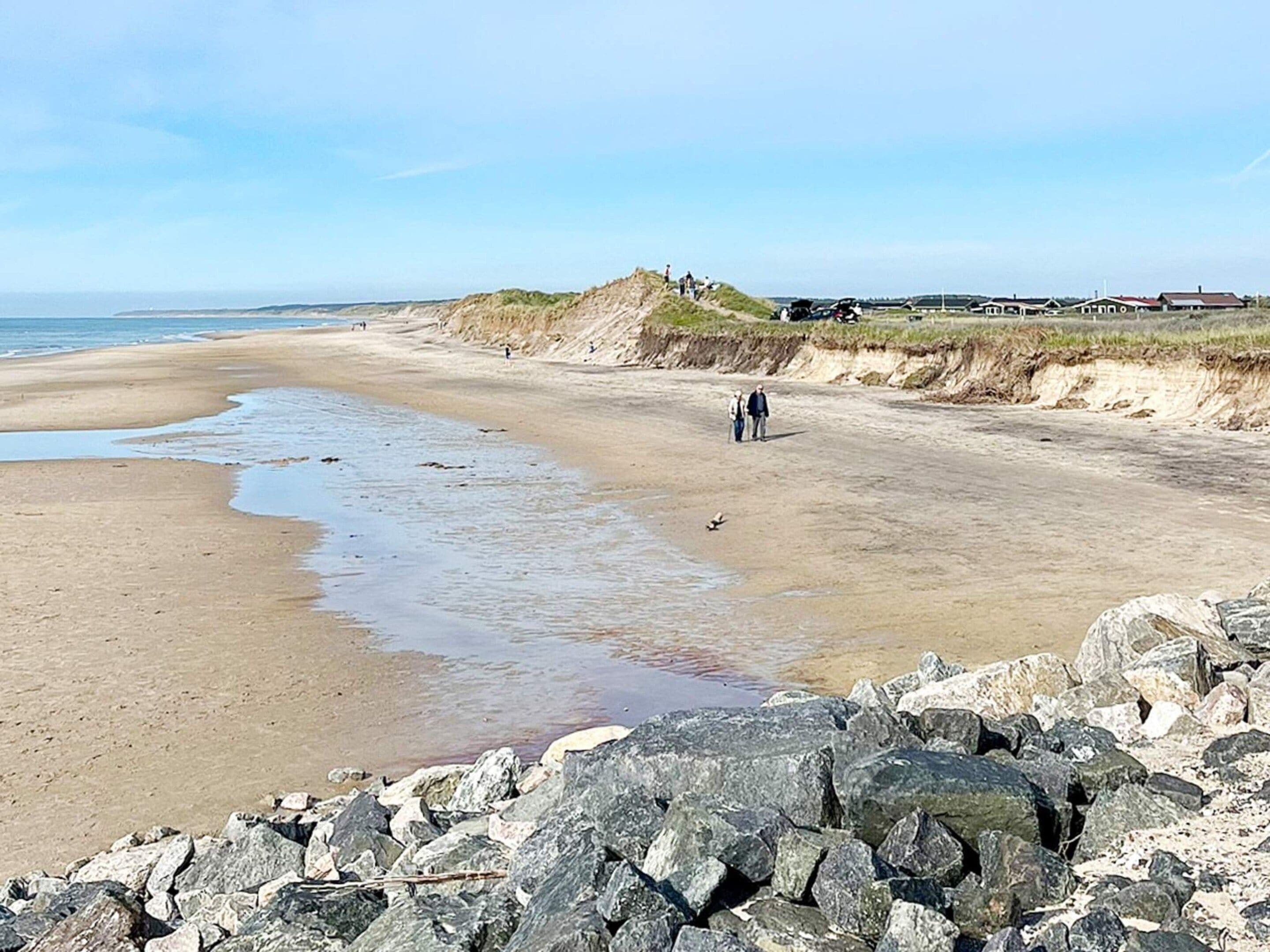 Uma praia com um primeiro plano rochoso, uma faixa de areia e algumas pessoas ao longe.