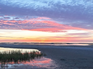 Uma praia com o pôr do sol, águas calmas e vegetação rala.