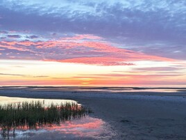 Uma praia com o pôr do sol, águas calmas e vegetação rala.