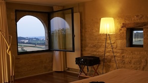 Quarto de hotel com uma grande janela em arco, uma luminária de pé, uma pequena mesa e uma cama.