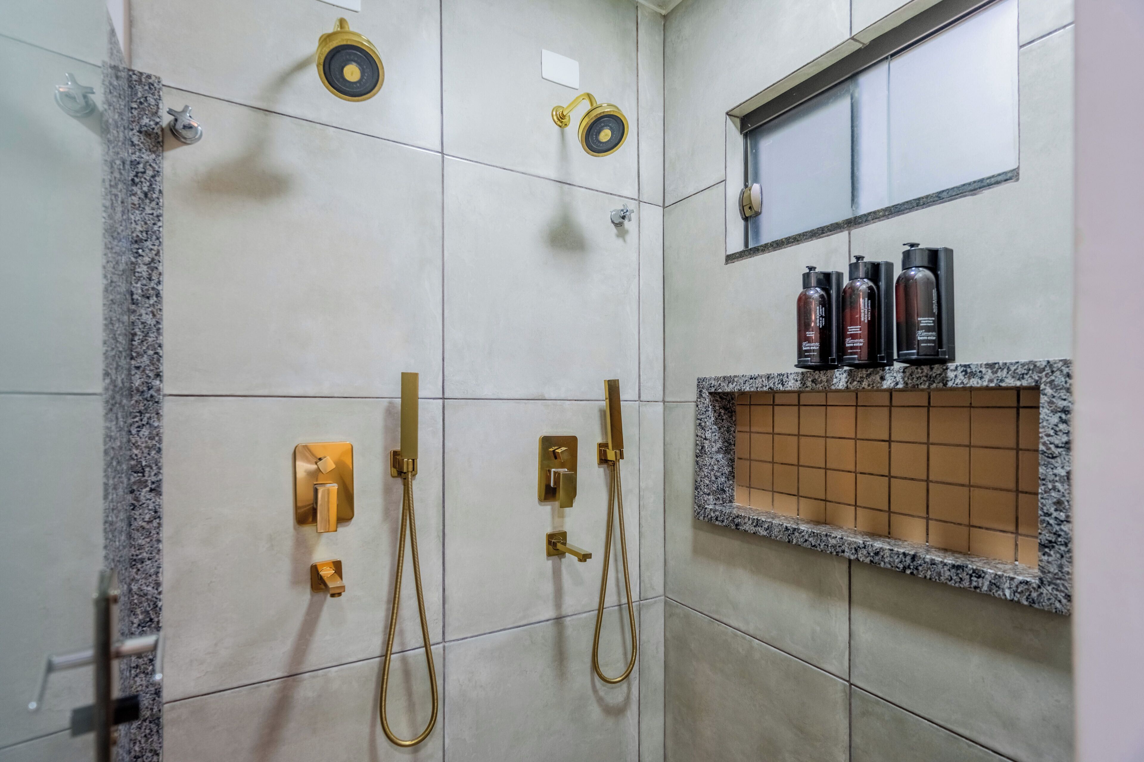 Área de chuveiro com detalhes dourados, uma janela e um prateleira com produtos de higiene pessoal.