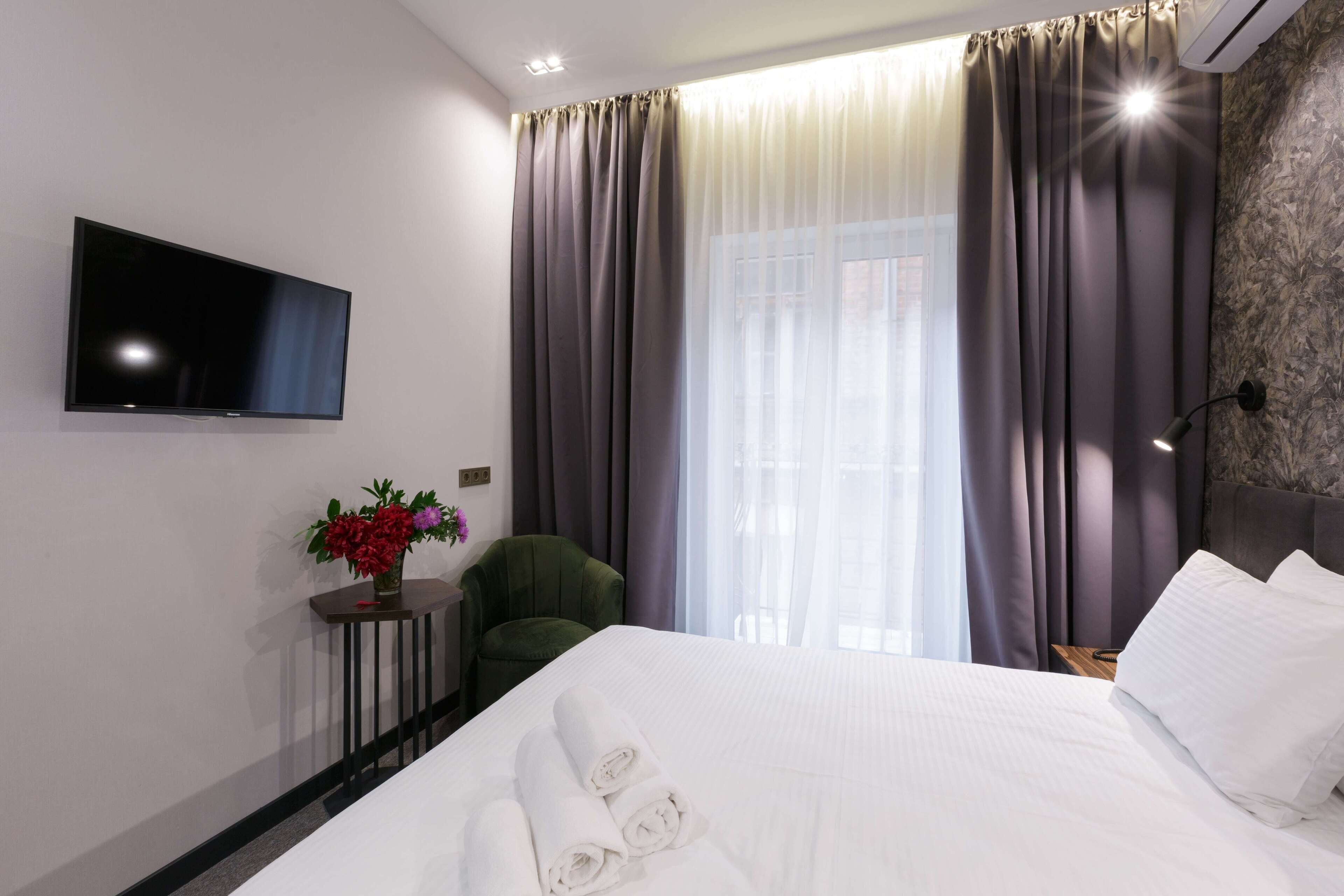 Quarto de hotel com uma cama grande, uma TV de tela plana na parede, uma mesinha de cabeceira com um vaso de flores e uma cadeira verde.