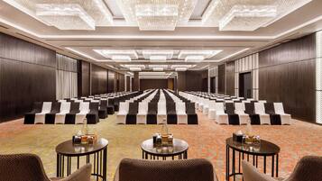 Um amplo hall de hotel com uma fileira longa de cadeiras preto e branco, uma área de estar central com mesas redondas e um candelabro com várias luminárias.