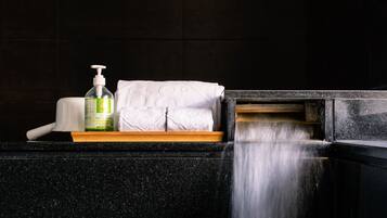 Uma bancada preta com uma embalagem de sabonete líquido, uma toalha branca e um tabuleiro de madeira.