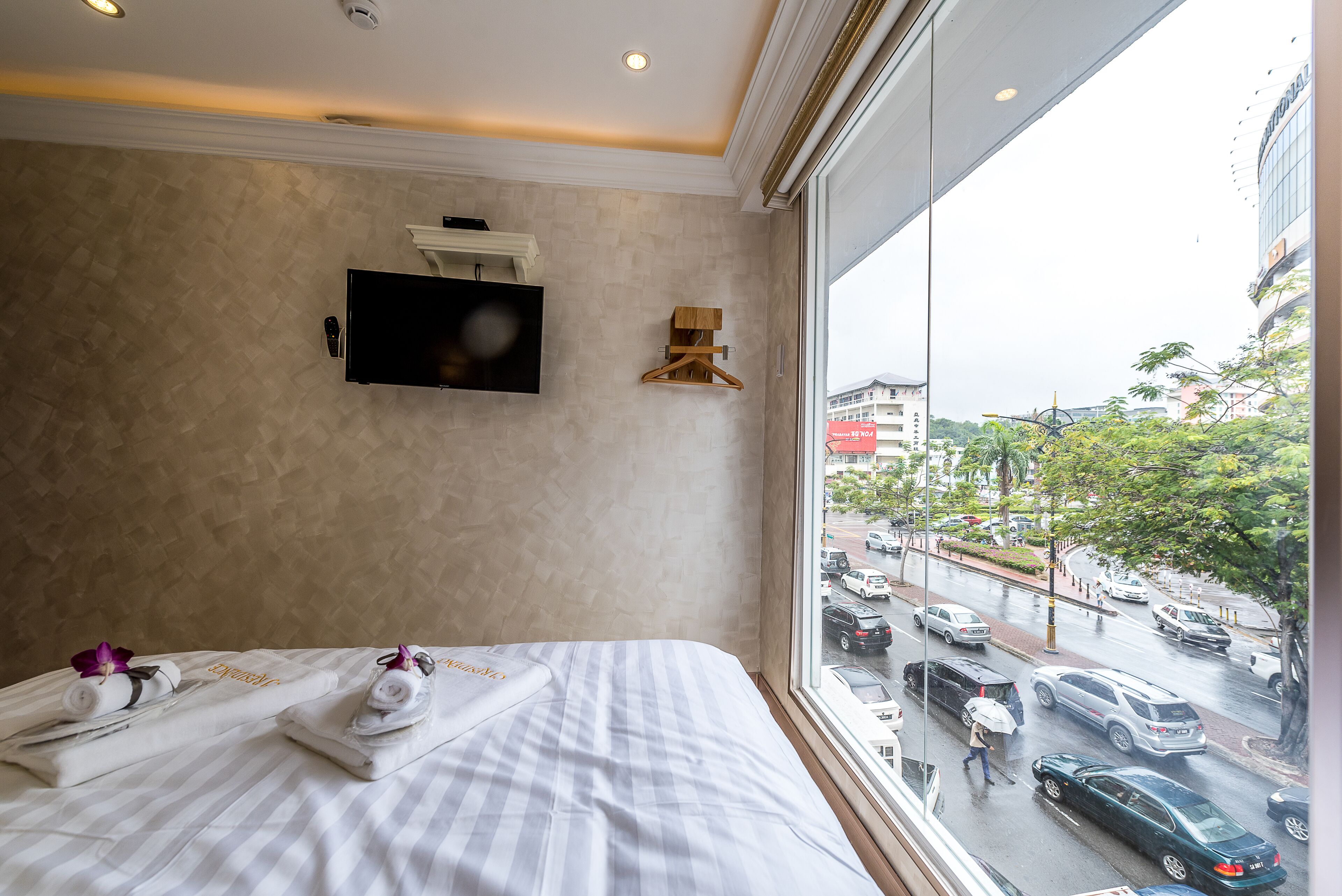 Quarto de hotel com uma cama, uma televisão fixada na parede e vista para a rua através de uma grande janela.