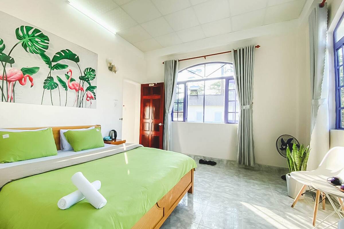 Quarto de hotel com cama, almofadas verdes, pintura de folhas tropicais e flamingos na parede, janela com cortinas e uma cadeira.