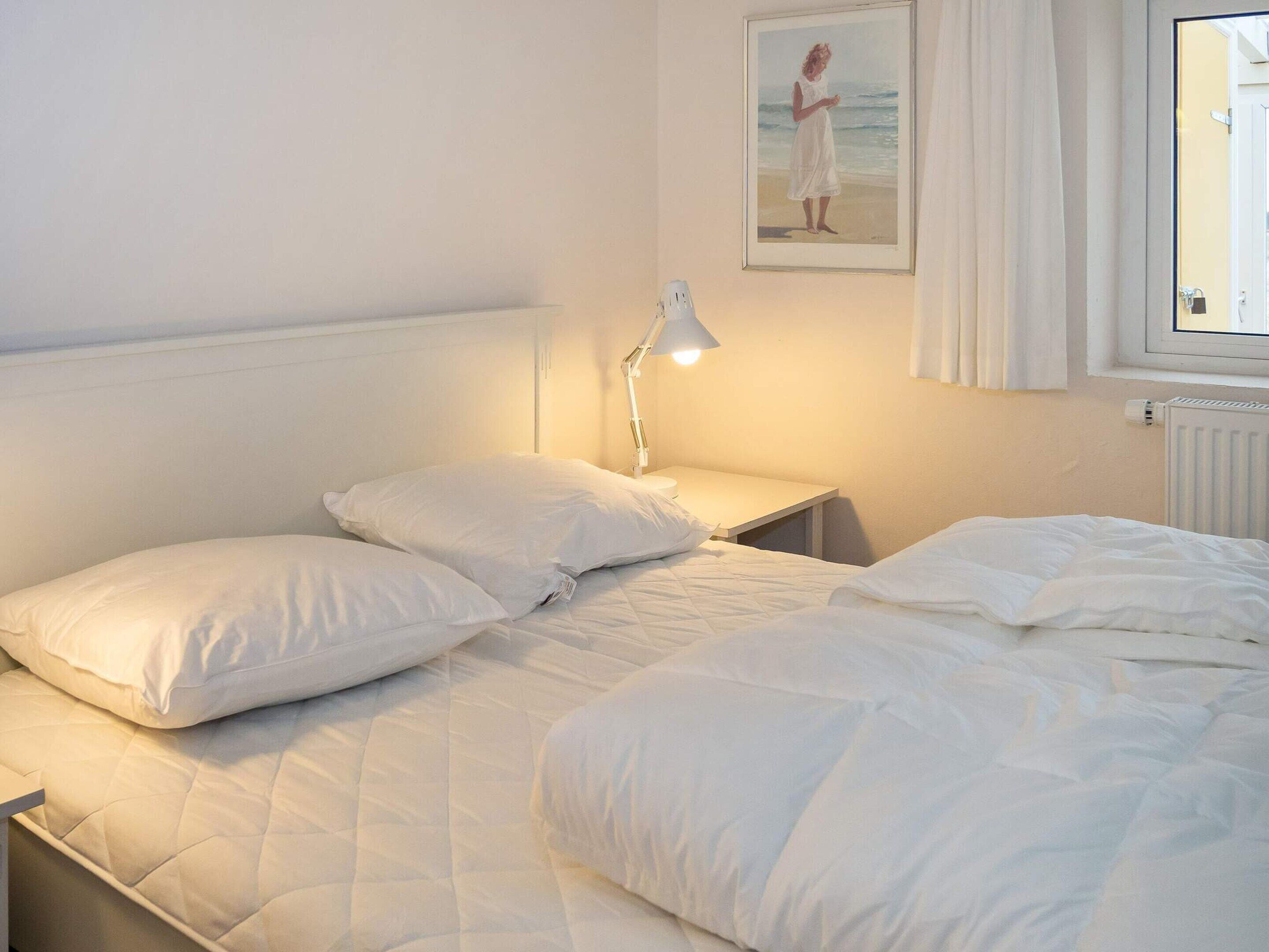 Quarto com cama, almofadas, mesa de cabeceira com abajur, janela com cortinas e um quadro na parede.