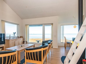 Ampla sala de estar com uma grande janela vista para a praia, mesa de jantar de madeira e assentos confortáveis.