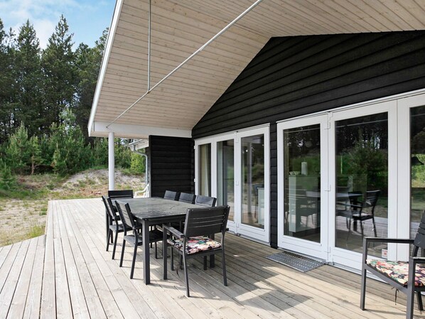 Um deck com mesa e cadeiras, área coberta com telhado inclinado e porta de vidro dando para um jardim.