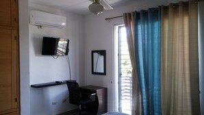 Quarto com escrivaninha, cadeira, televisão, aparelho de ar condicionado e janela com cortinas.