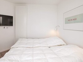 Quarto minimalista com cama, televisão fixada na parede, luminária e quadro emoldurado.
