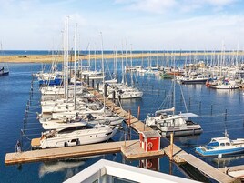 Uma marina com vários barcos atracados, um pequeno edifício vermelho e uma praia de areia ao fundo.
