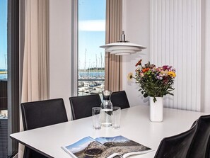 Uma sala de jantar com uma mesa posta para dois, um vaso de flores e vista para um porto através de grandes janelas.