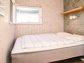 Uma cama de solteiro com colcha e almofadas listradas, uma janela acima e uma parede de tijolos ao fundo.