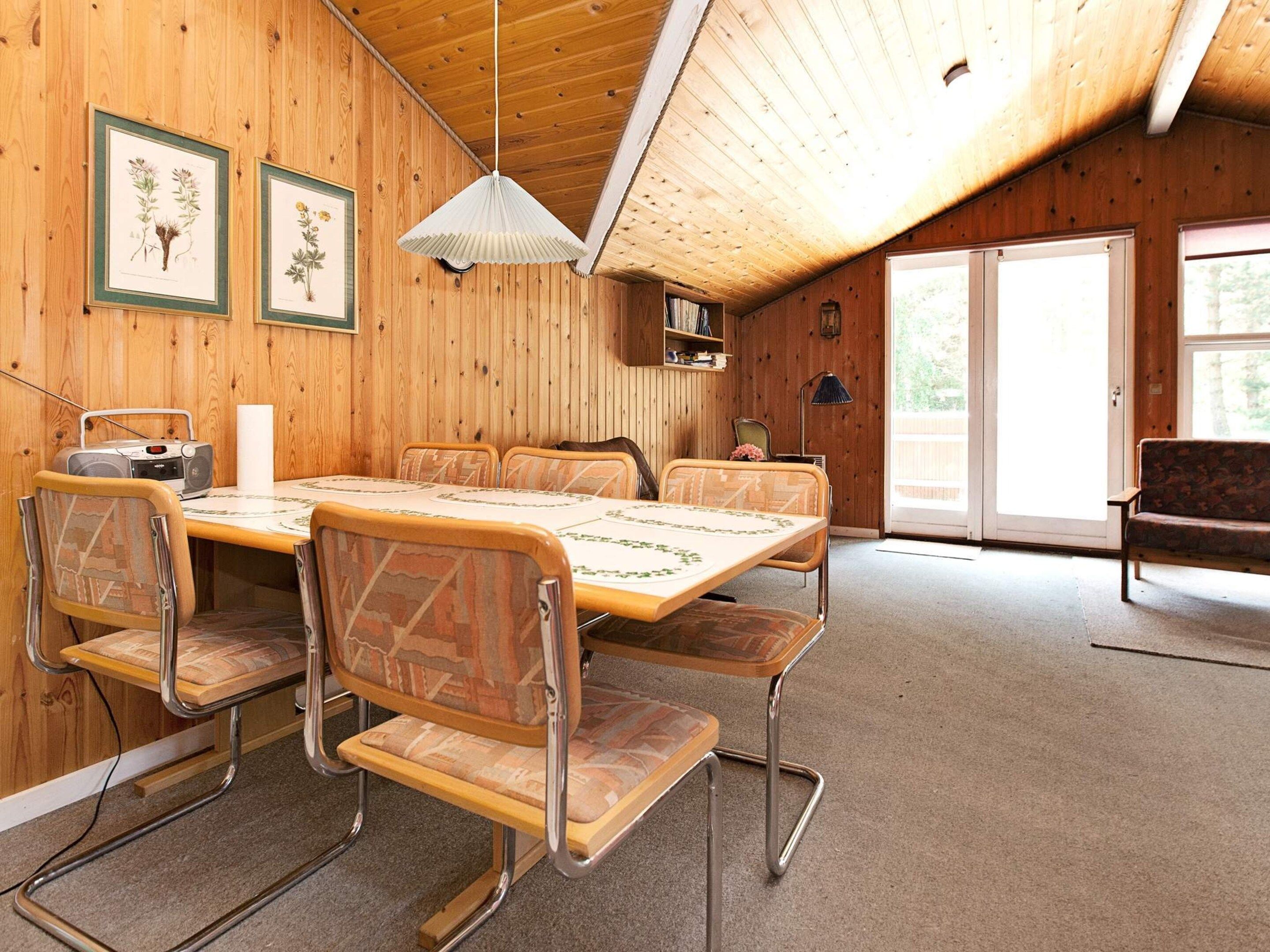 Um quarto com painéis de madeira, uma mesa de jantar, cadeiras, um rádio, ilustrações botânicas emolduradas e um banco.