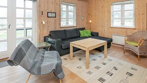Um quarto com painéis de madeira, um sofá, uma mesa, uma cadeira de vime e janelas amplas.
