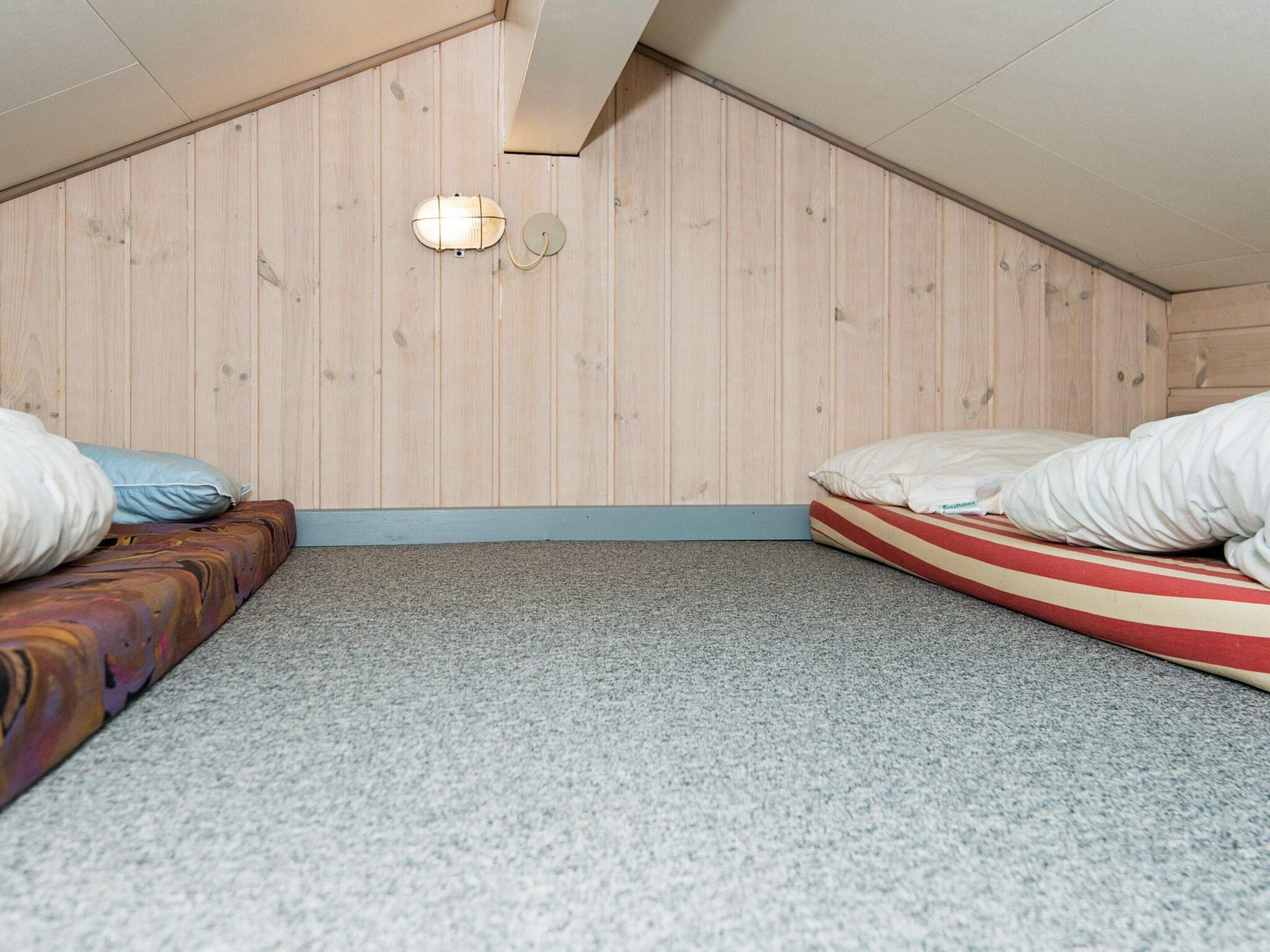 Quarto pequeno com teto inclinado, duas camas, luminária na parede e piso coberto por carpete.