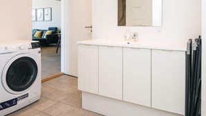 Uma lavanderia com máquina de lavar, pia com espelho e porta para uma sala de estar com sofá e mesa de centro.