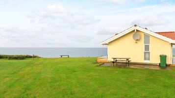 Um edifício amarelo com telhado vermelho, um banco e uma mesa de piquenique em uma área gramada com vista para o mar.