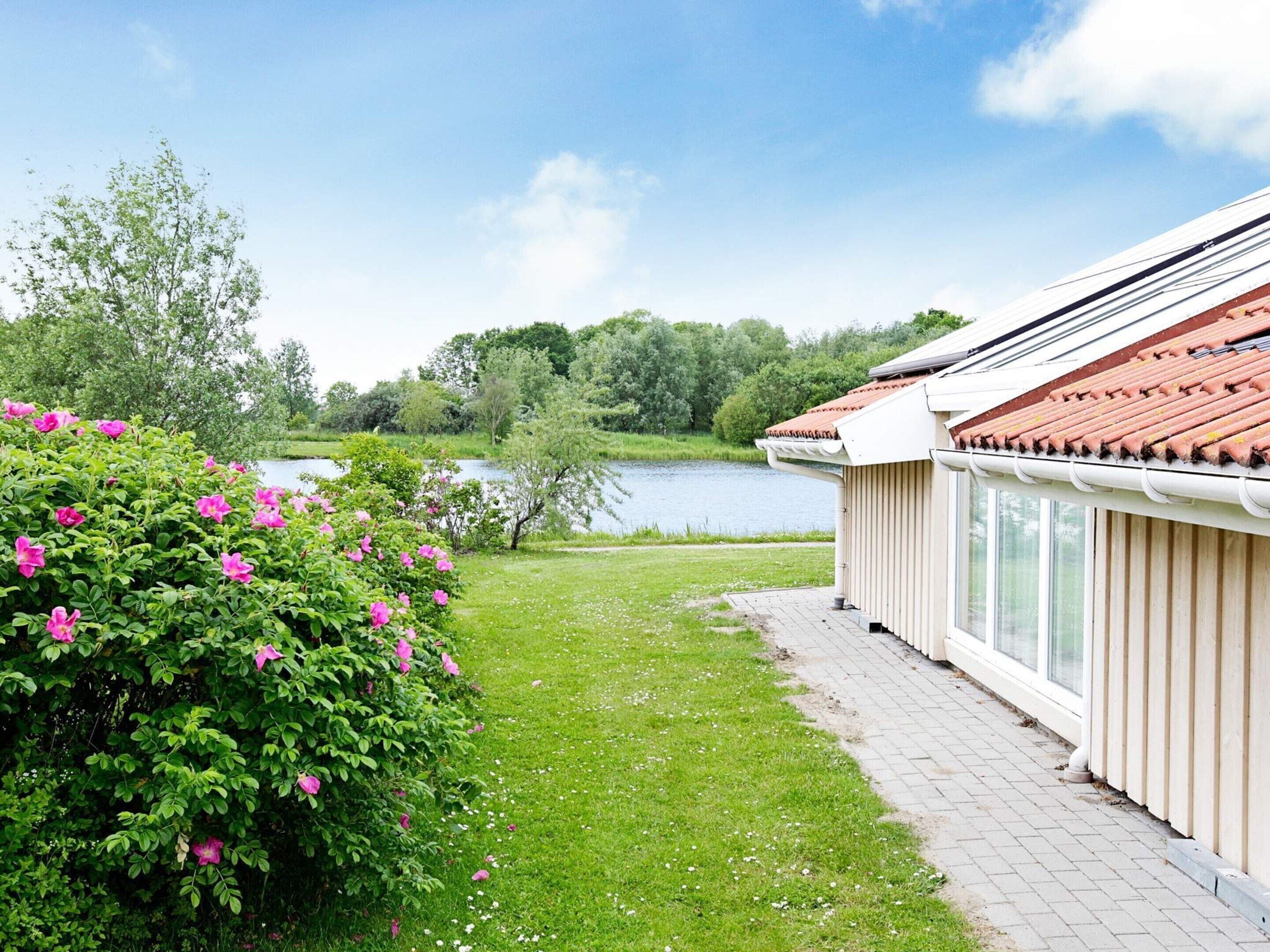 Um edifício à beira do lago, com um calçadão pavimentado, um arbusto com flores cor-de-rosa e vista para o lago.