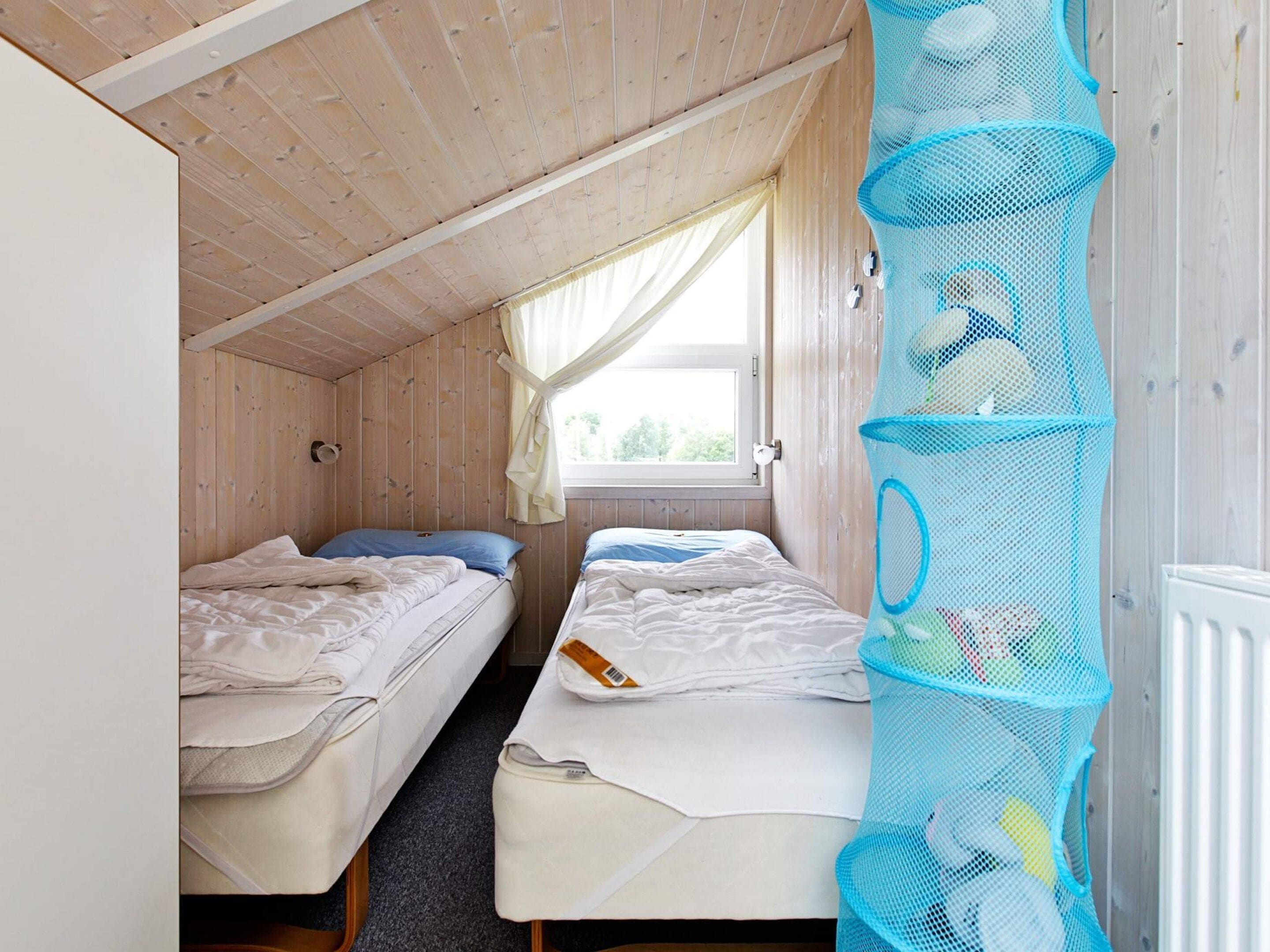 Quarto pequeno e acolhedor com duas camas de solteiro, uma janela com cortinas e um cesto azul pendurado.