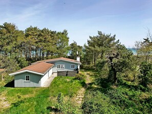 Uma casa térrea com telhado inclinado, cercada por árvores densas e um céu claro.
