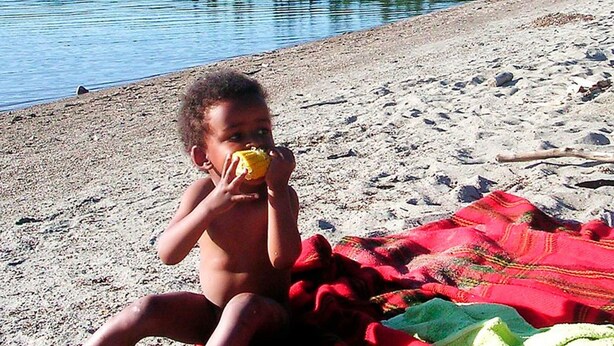 Criança sentada na praia arenosa, com um corpo d'água e árvores ao fundo.