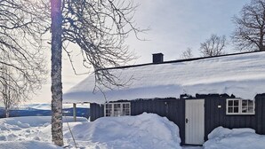 Uma cabana de madeira rodeada por neve, com céu claro e árvores secas ao fundo.