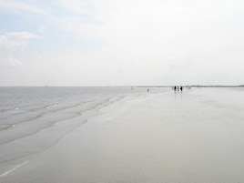 Uma extensa praia de areia com ondas suaves, céu claro e um litoral distante.