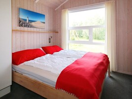 Quarto com cama de madeira, roupa de cama vermelha e branca, um quadro na parede e uma janela com cortinas.