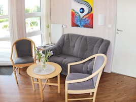 Quarto com um sofá cinzento, duas cadeiras de madeira, uma mesa redonda pequena com um vaso de flores e um quadro na parede.