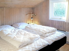 Quarto com paredes de madeira, cama com roupa de cama branca, luminária de cabeceira e janela com vista para árvores.