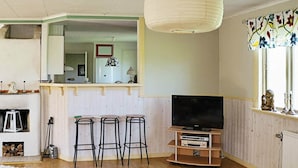 Uma sala de estar com piso de madeira, uma TV sobre um suporte de madeira, três banquetas, um tapete branco e fofo, e uma janela com cortina floral.
