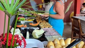 Uma mulher servindo comida em um buffet ao ar livre, com diversos pratos e uma churrasqueira em primeiro plano.