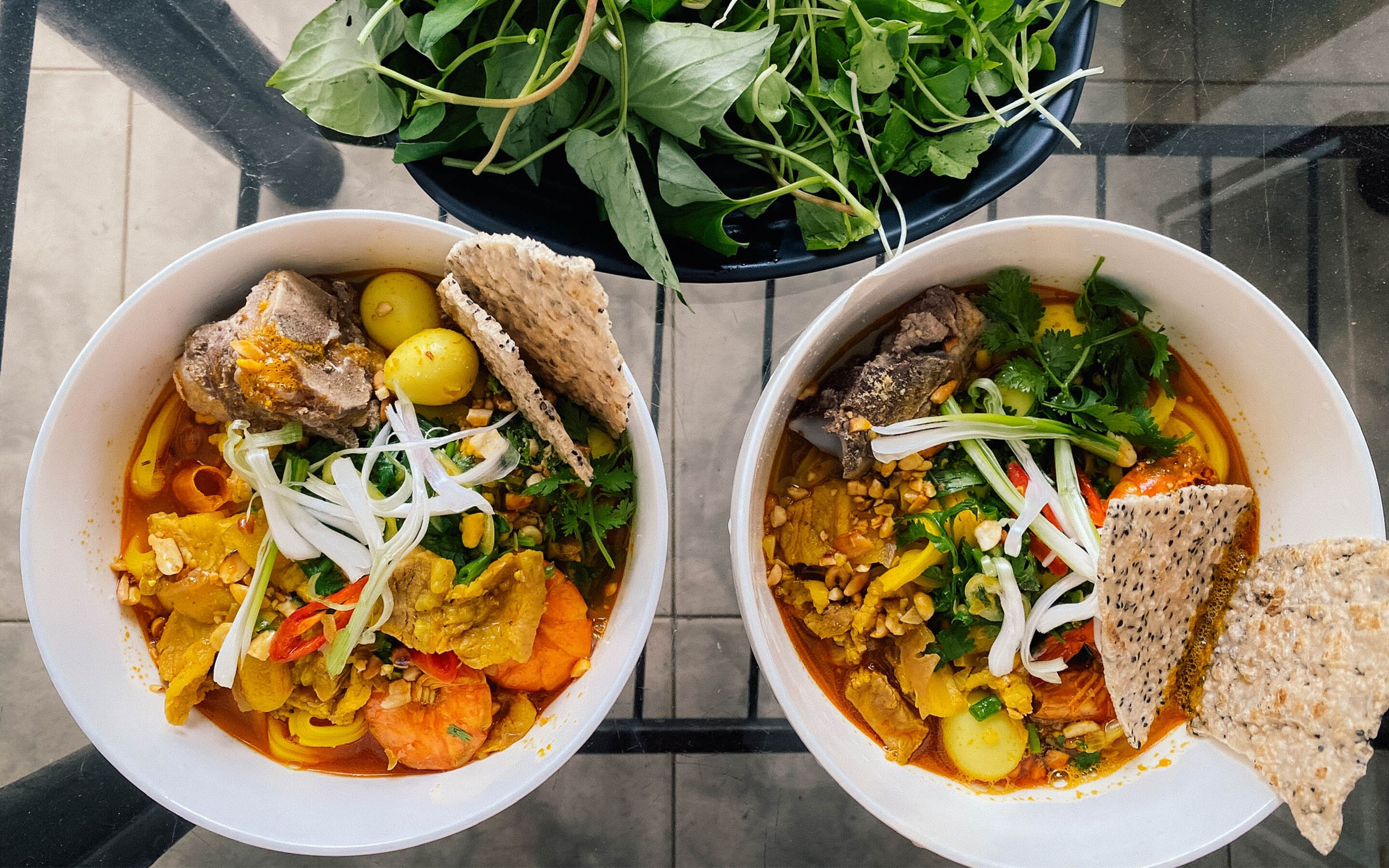 Duas tigelas de sopa com diversos ingredientes, como carne, legumes e ervas, servidas sobre uma mesa de vidro.