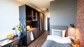 Quarto de hotel compacto com cozinha compacta, cama com cabeceira cinza, escrivaninha com cadeira e mesa de jantar com uma fruteira.