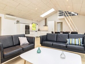Uma sala de estar moderna com dois sofás de couro preto, uma mesa de centro branca e uma área de cozinha com armários brancos e um forno embutido.