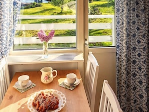 Quarto com mesa de madeira, duas cadeiras, janela com vista para uma paisagem verde e um vaso com flores.
