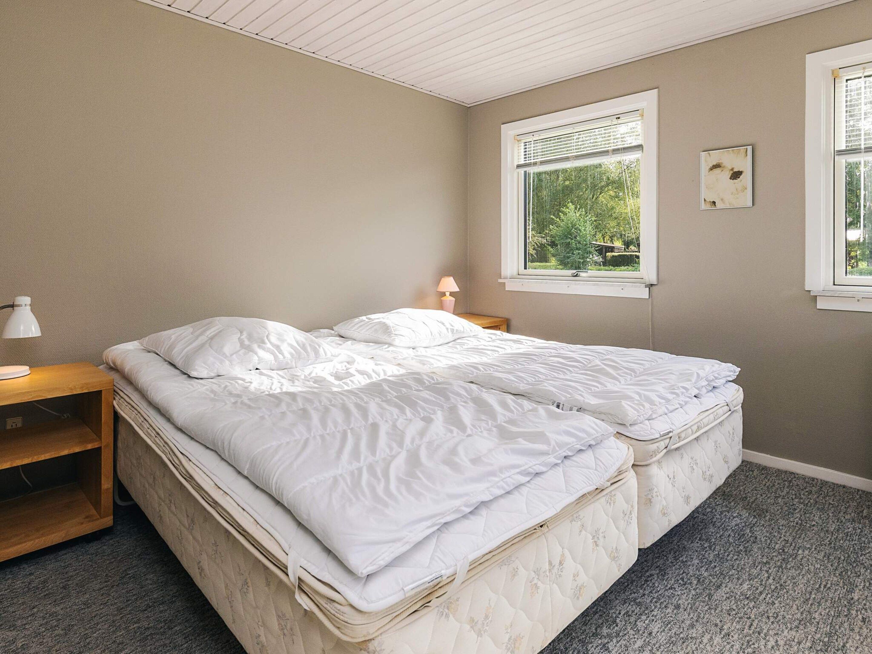 Quarto com cama de casal, mesa de cabeceira com luminária, janela com persianas e um quadro na parede.